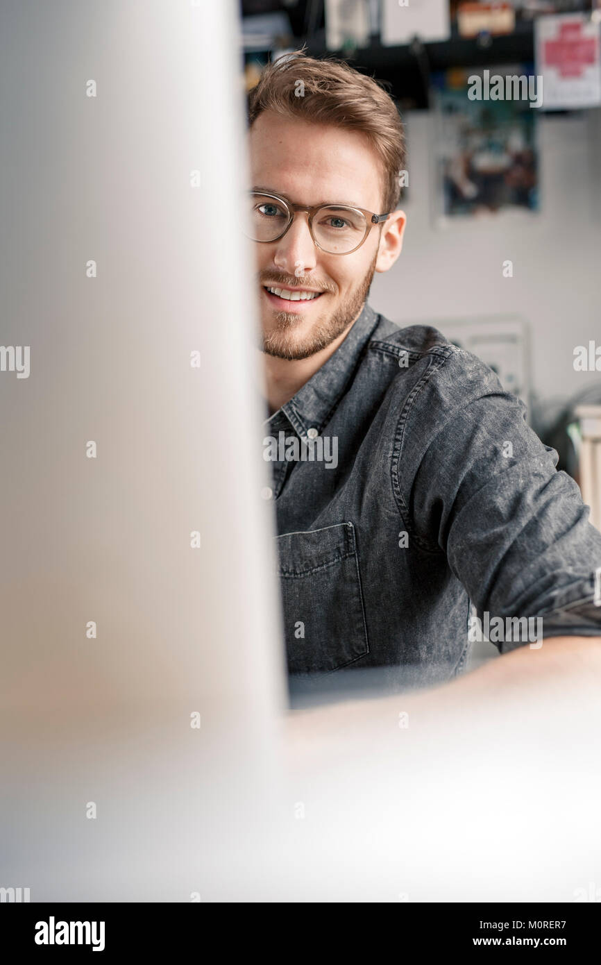 Portrait of smiling young man behind computer screen at desk at home ...