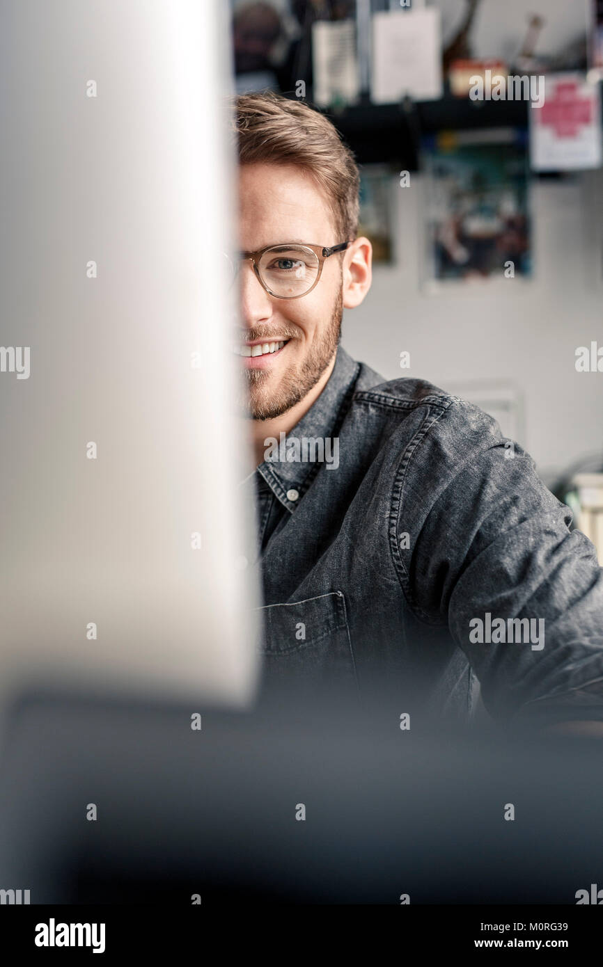 Portrait of smiling young man behind computer screen at desk at home ...