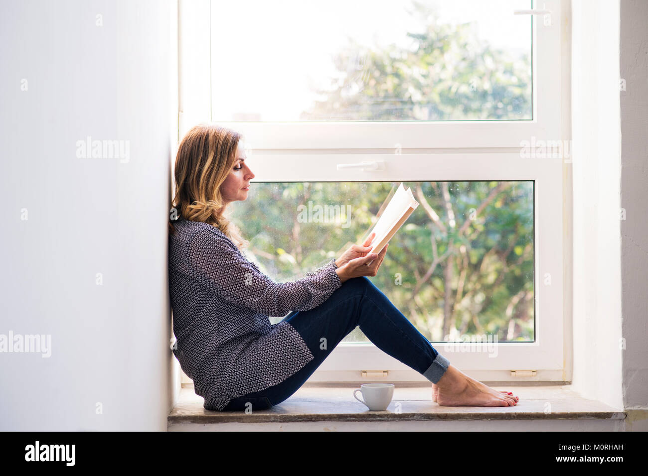 Woman sitting at home on the window sill, reading a book Stock Photo ...