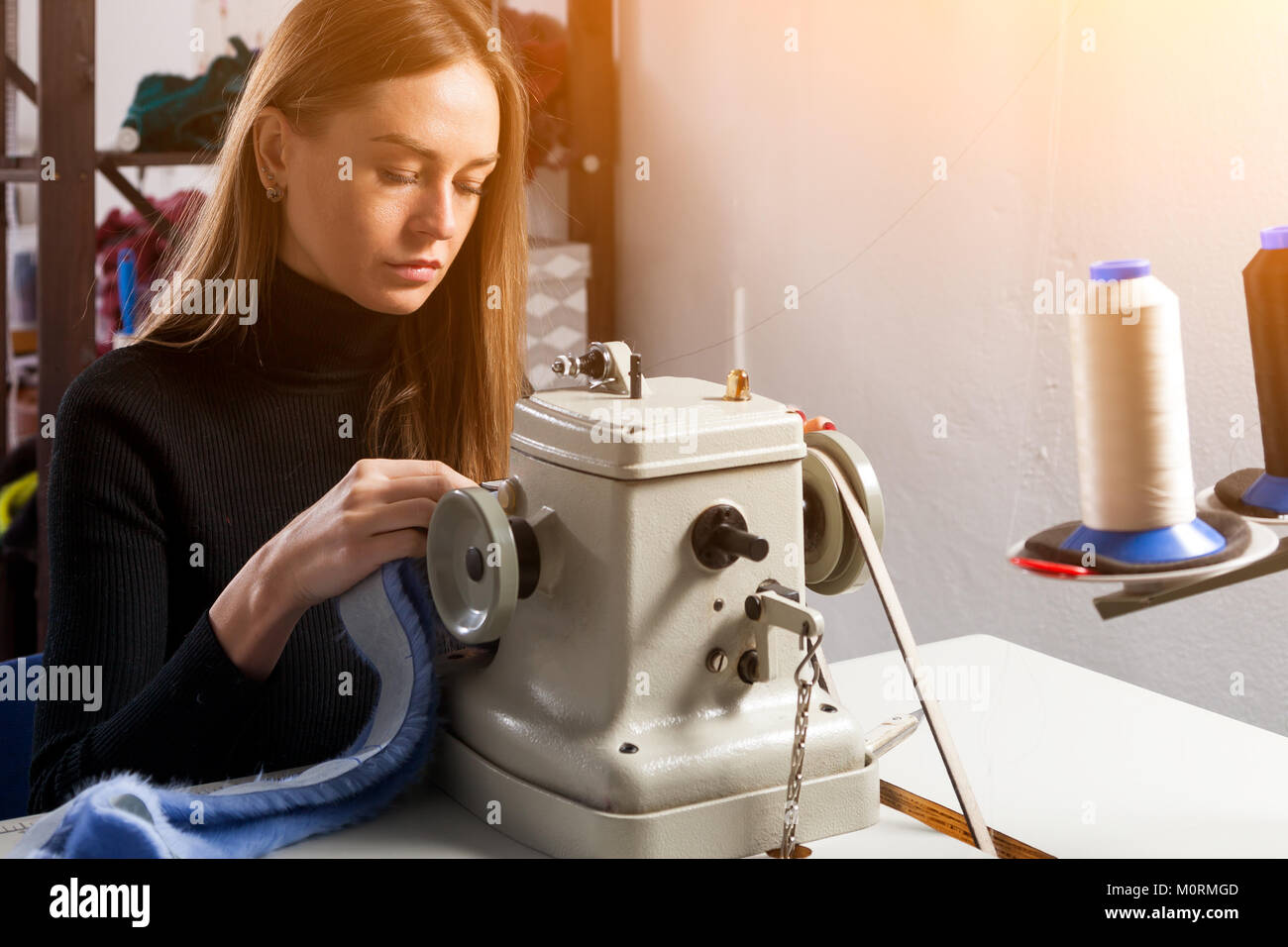 Young woman Furrier sewing blue fur coat on sewing machine in workshop ...