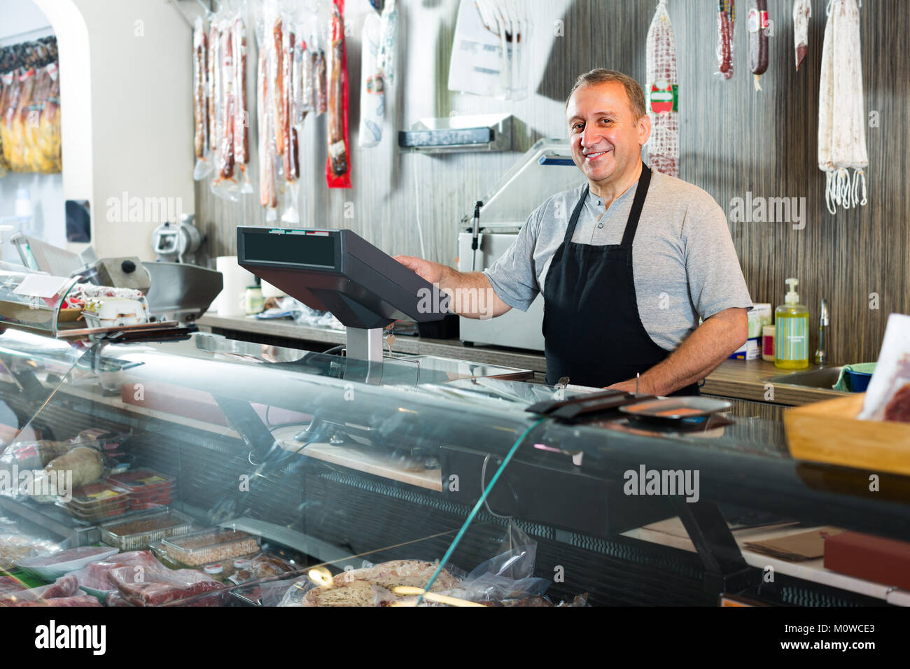 Mature male butcher with assortment of wurst and smoked meat at counter ...
