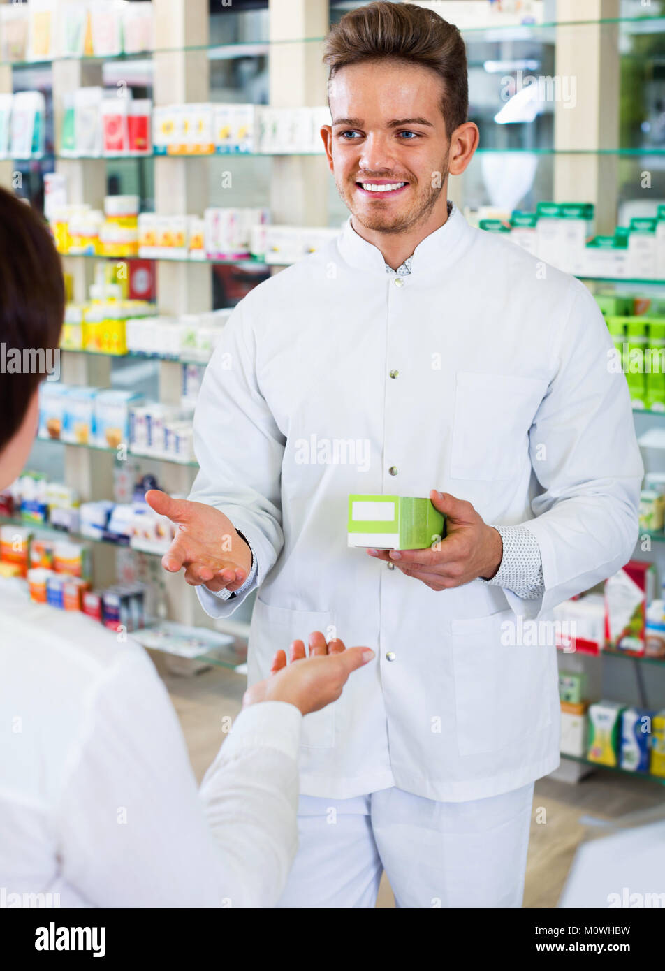 Positive man pharmacist wearing uniform assisting customers in ...