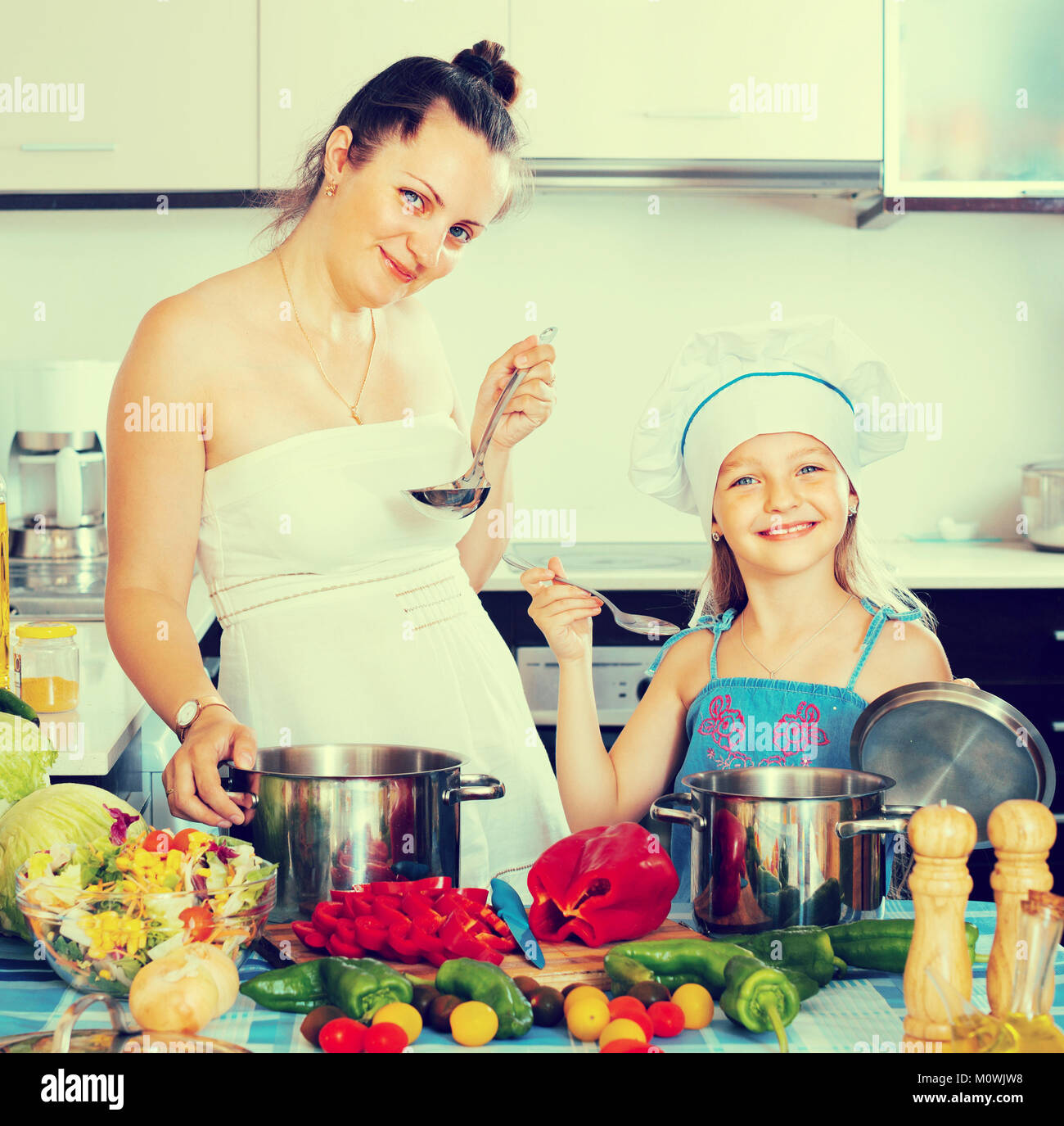 Smiling little girl and her mom cooking vegetable soup at home kitchen ...