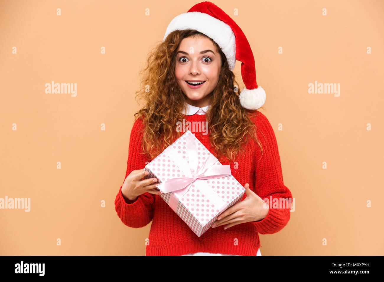 Portrait of a happy cheery girl dressed in santa hat holding gift box ...