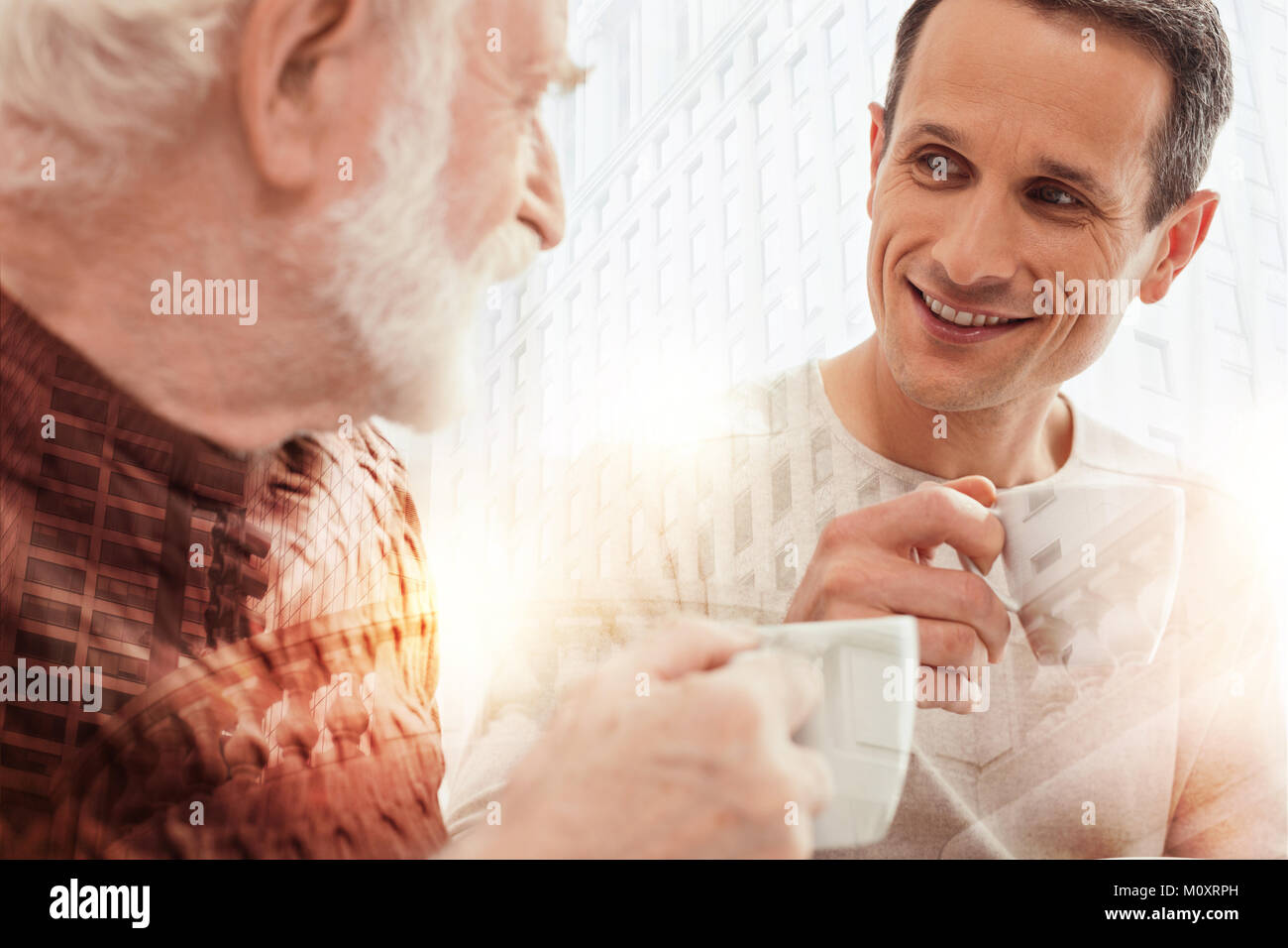 Kind attentive man looking at his aged father while drinking coffee ...