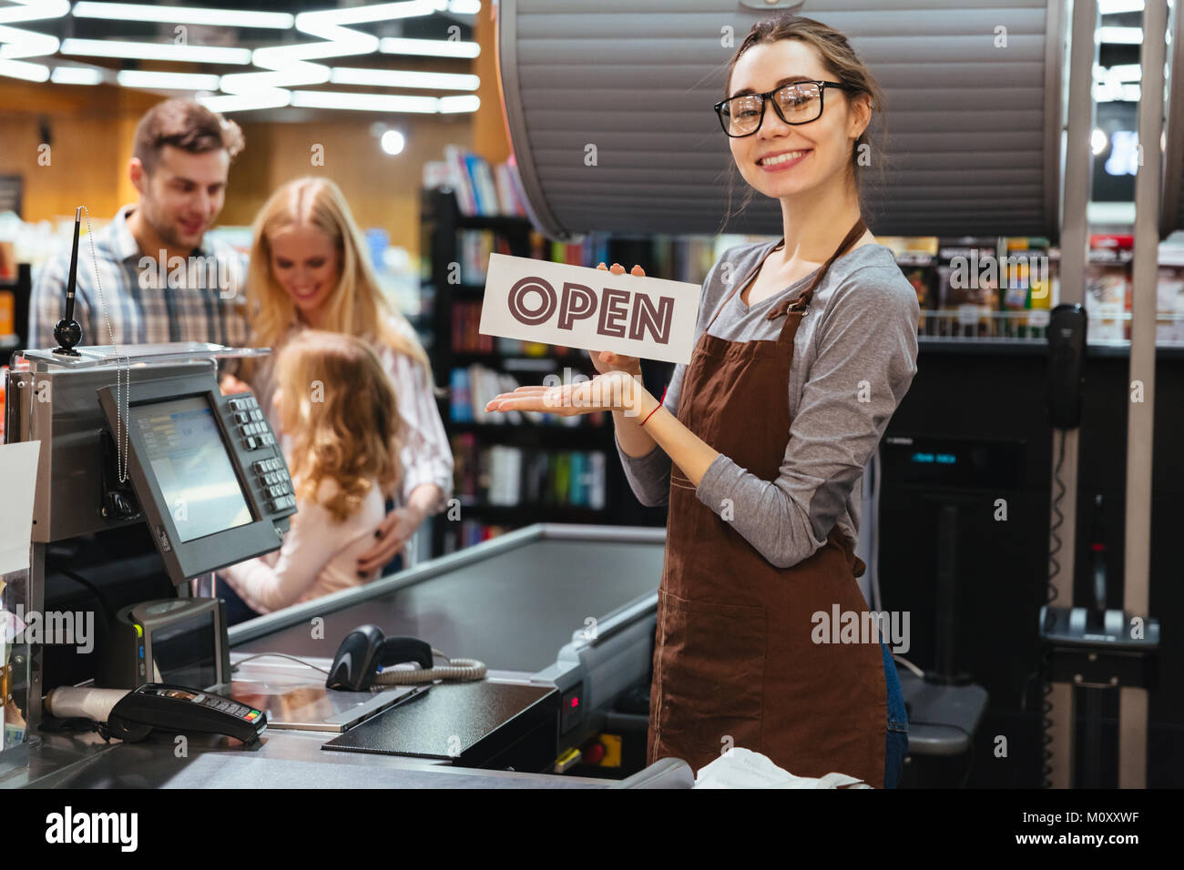 Portrait of happy woman cashier holding open sign while standing at ...