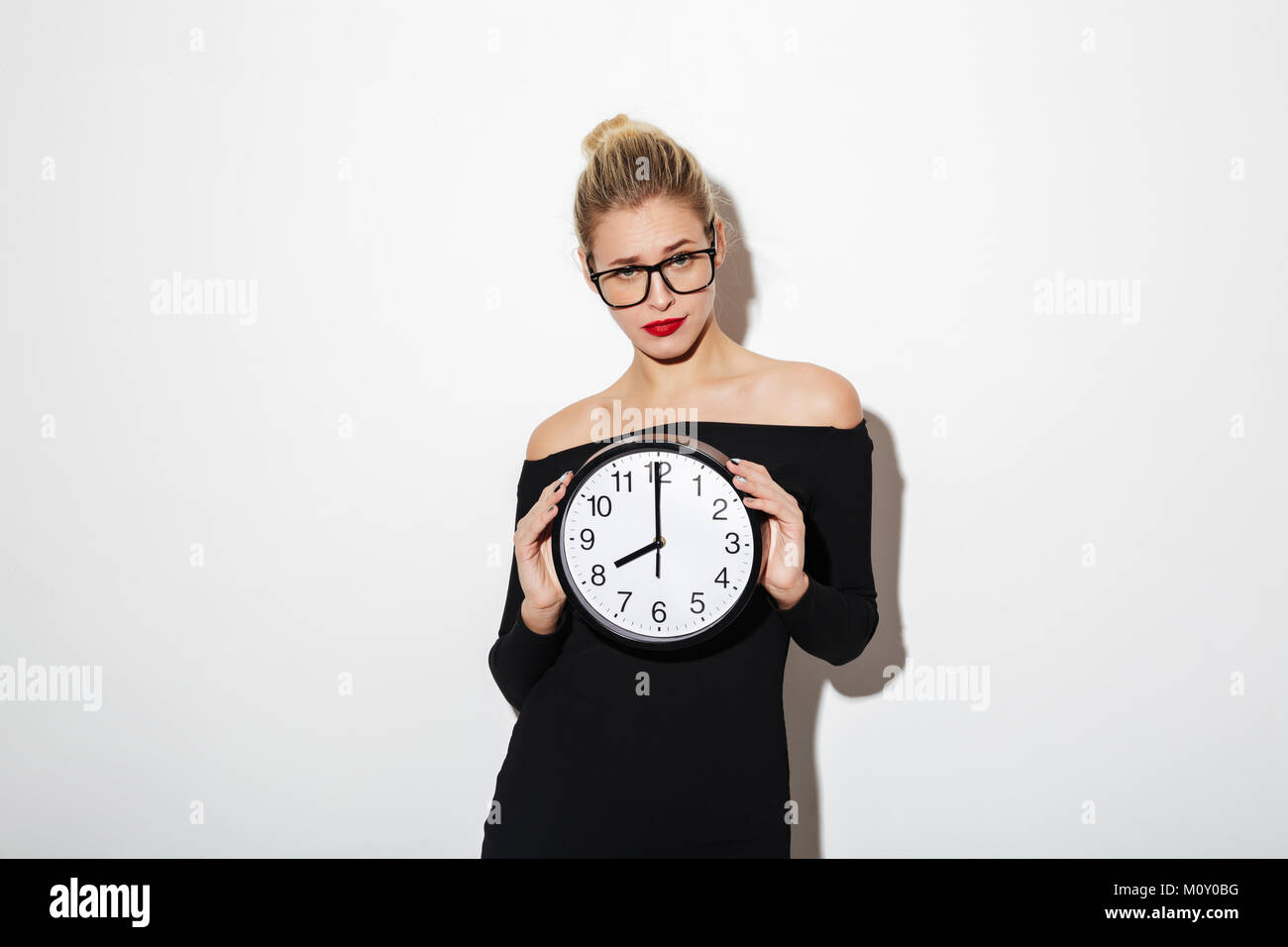 Confused business woman in dress and eyeglasses holding clock while ...