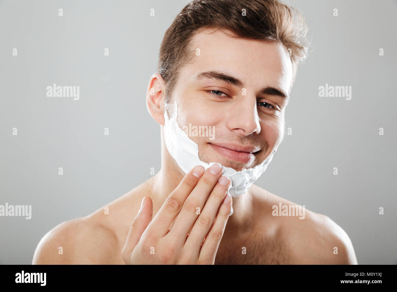 Close up portrait of a happy man applying shaving foam on his face isolated over gray background ...
