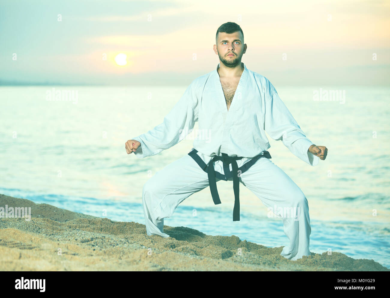 Young man is training the Shiko-dachi stance on the beach near the sea ...