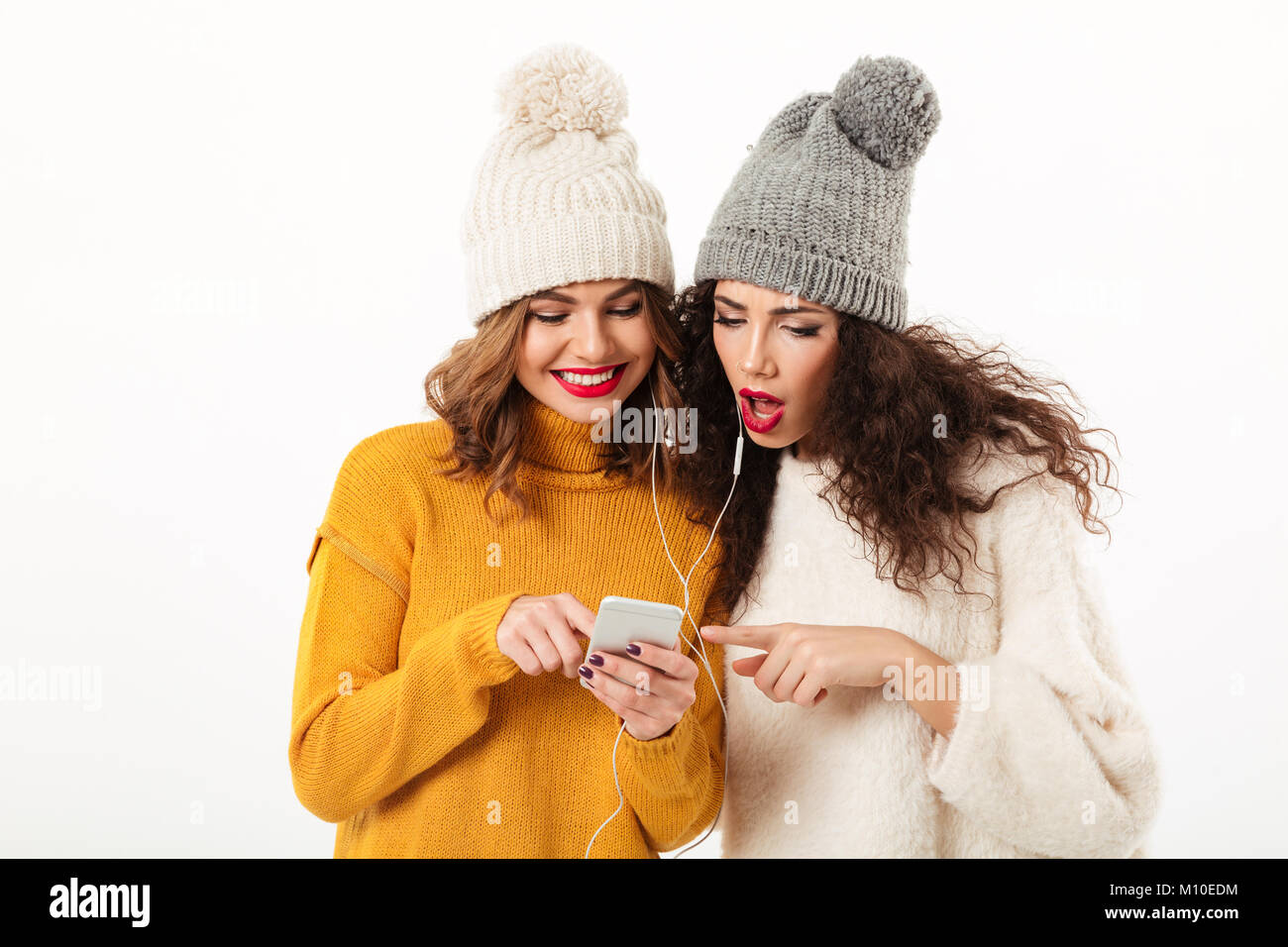 Two beauty girls in sweaters and hats standing together while using ...