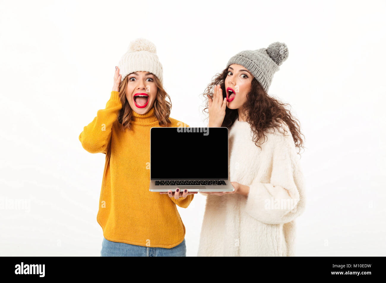 Two shocked girls in sweaters and hats standing together while showing ...