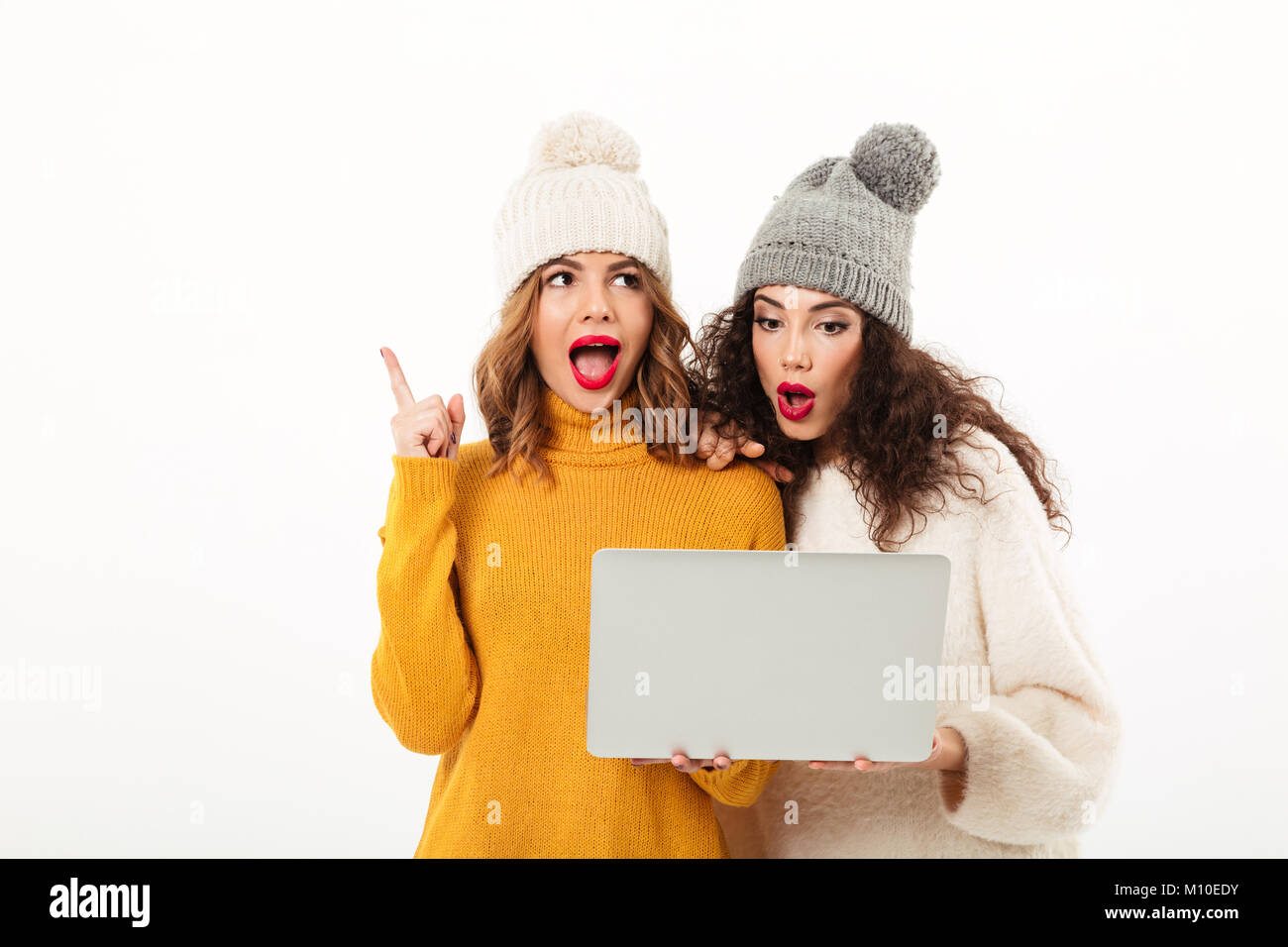Two surprised girls in sweaters and hats standing together while using ...
