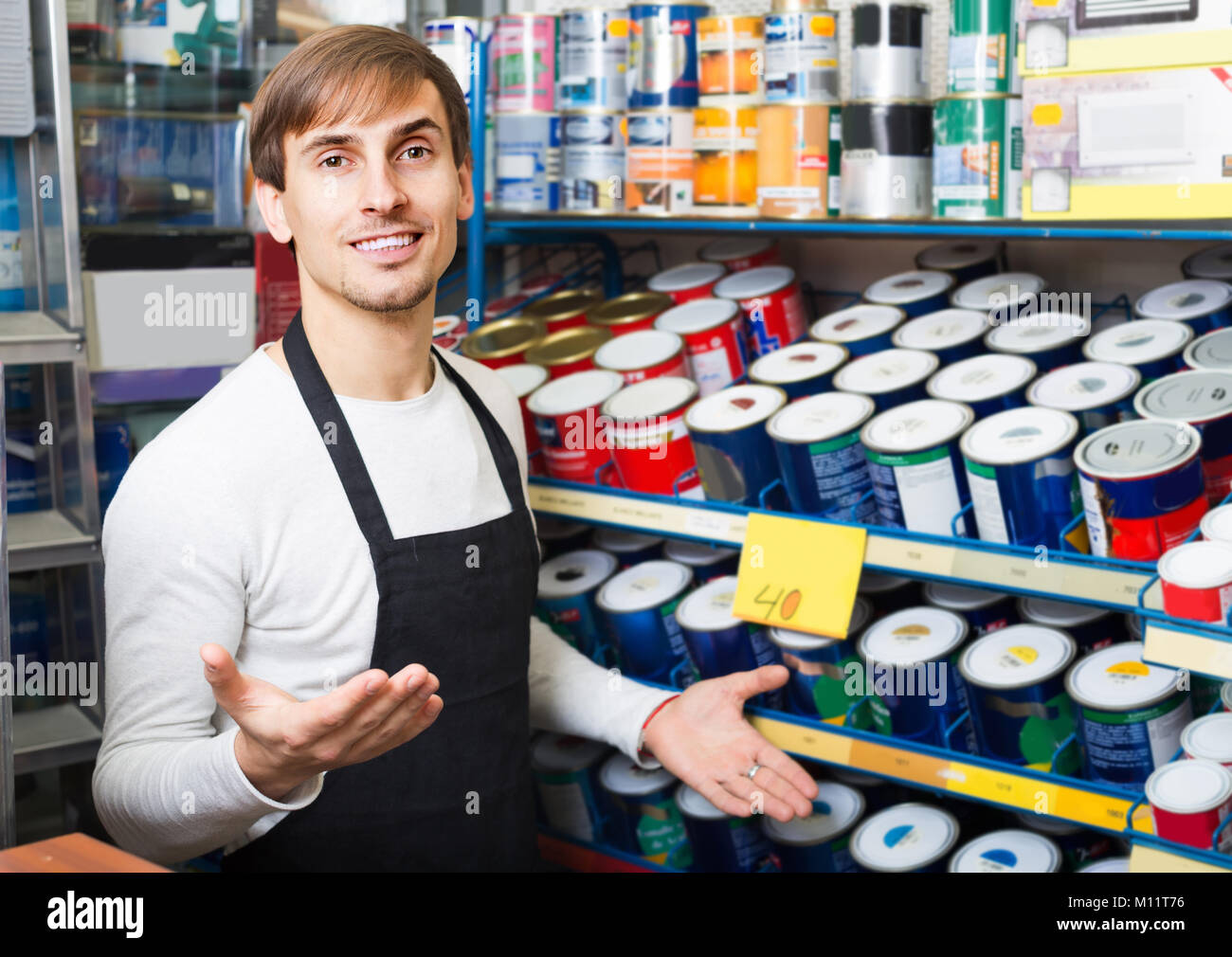 Friendly salesman in apron near shelves with paint and enamel in ...