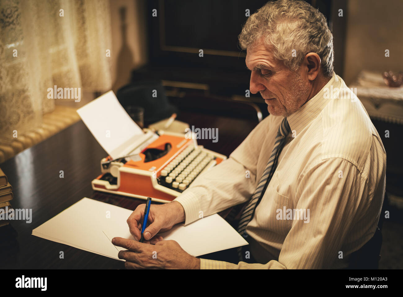 Retro senior man writer is writing with pencil beside obsolete ...