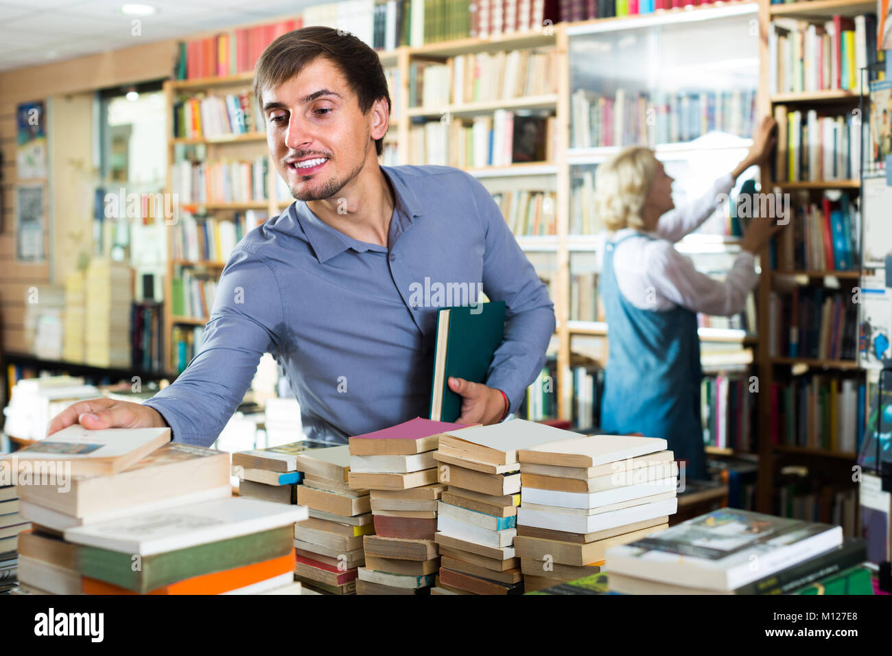 portrait of young positive man standing among bookshelves and searching ...