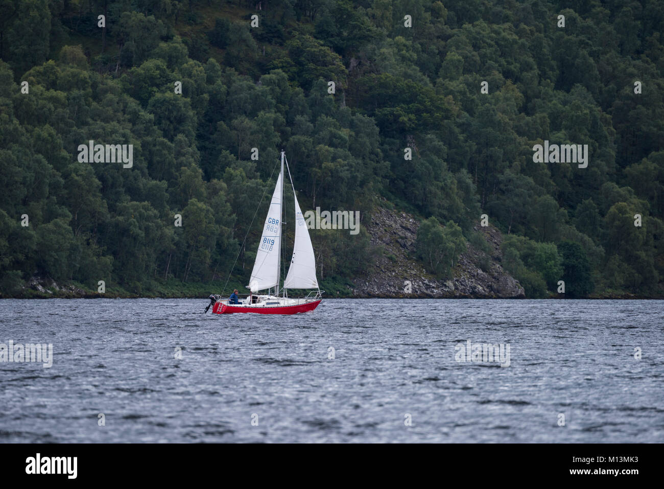 With steep wooded hillside beyond, a small red boat with 2 sails, is ...