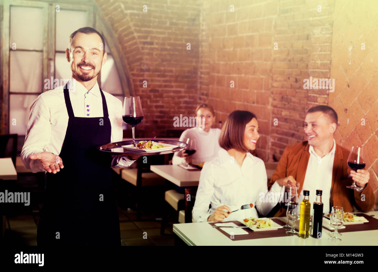 Positive male waiter welcoming guests to country restaurant Stock Photo ...