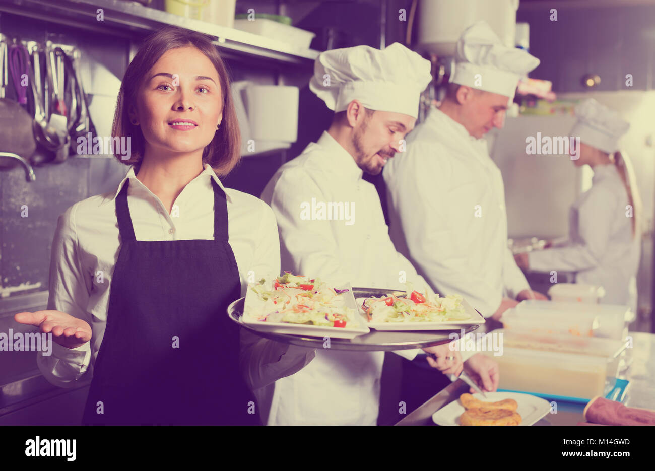 Laughing female waiter taking ordered dishes from restaurant’s kitchen ...