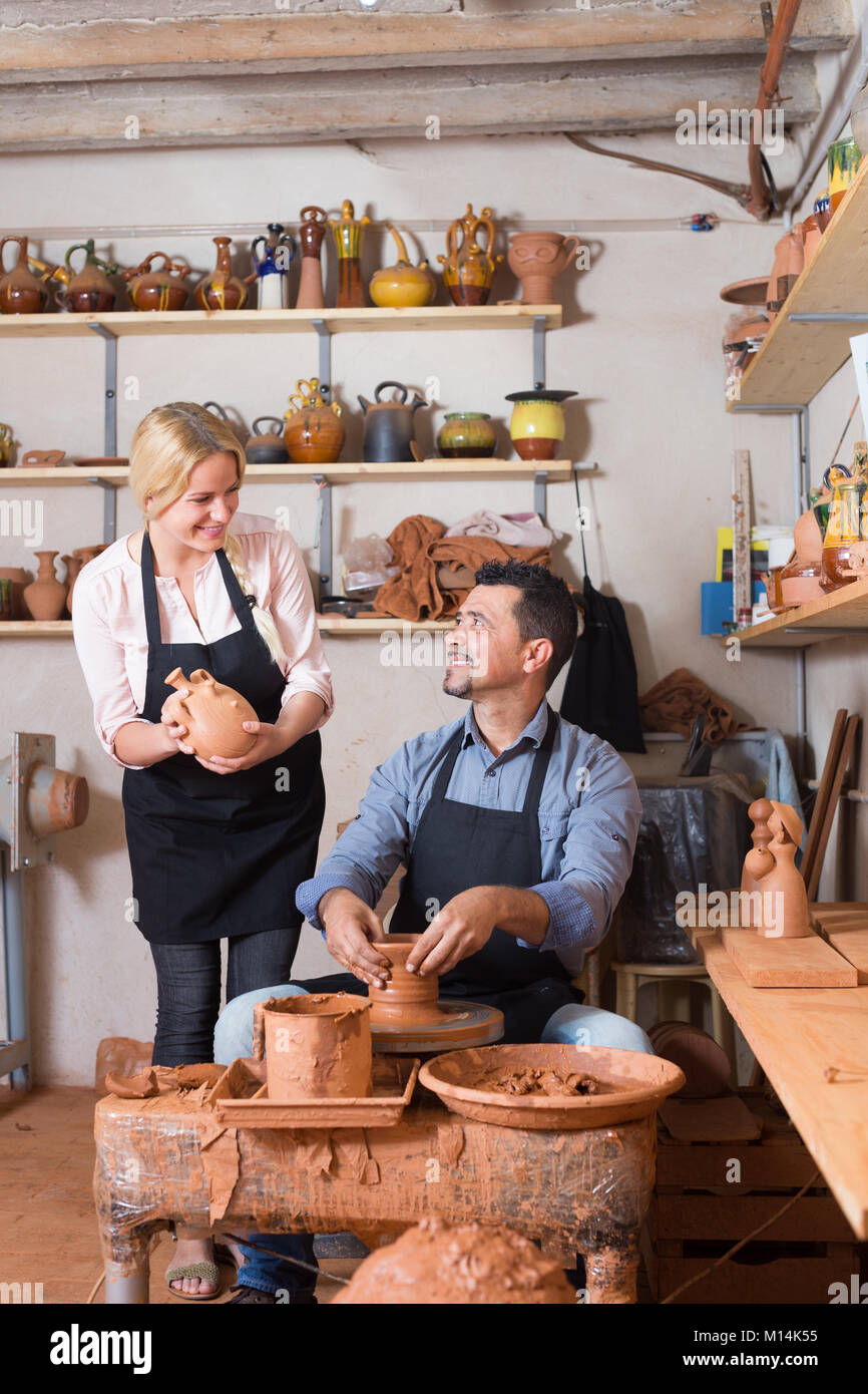 Positive craftsman making pot using pottery wheel and talking to woman ...