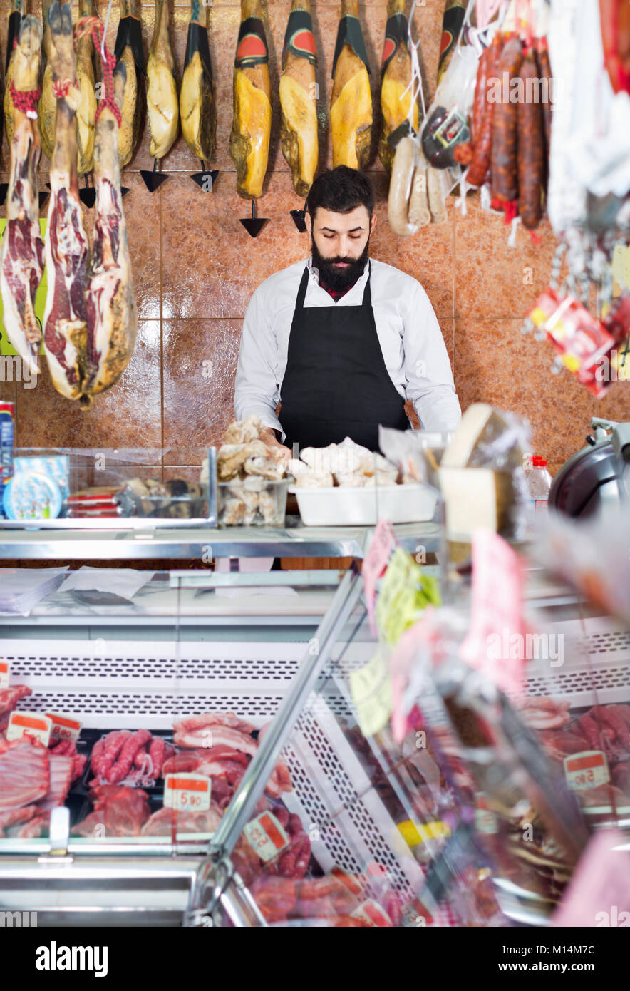 Cheerful positive man seller grouping meat to sell in butcher’s shop ...