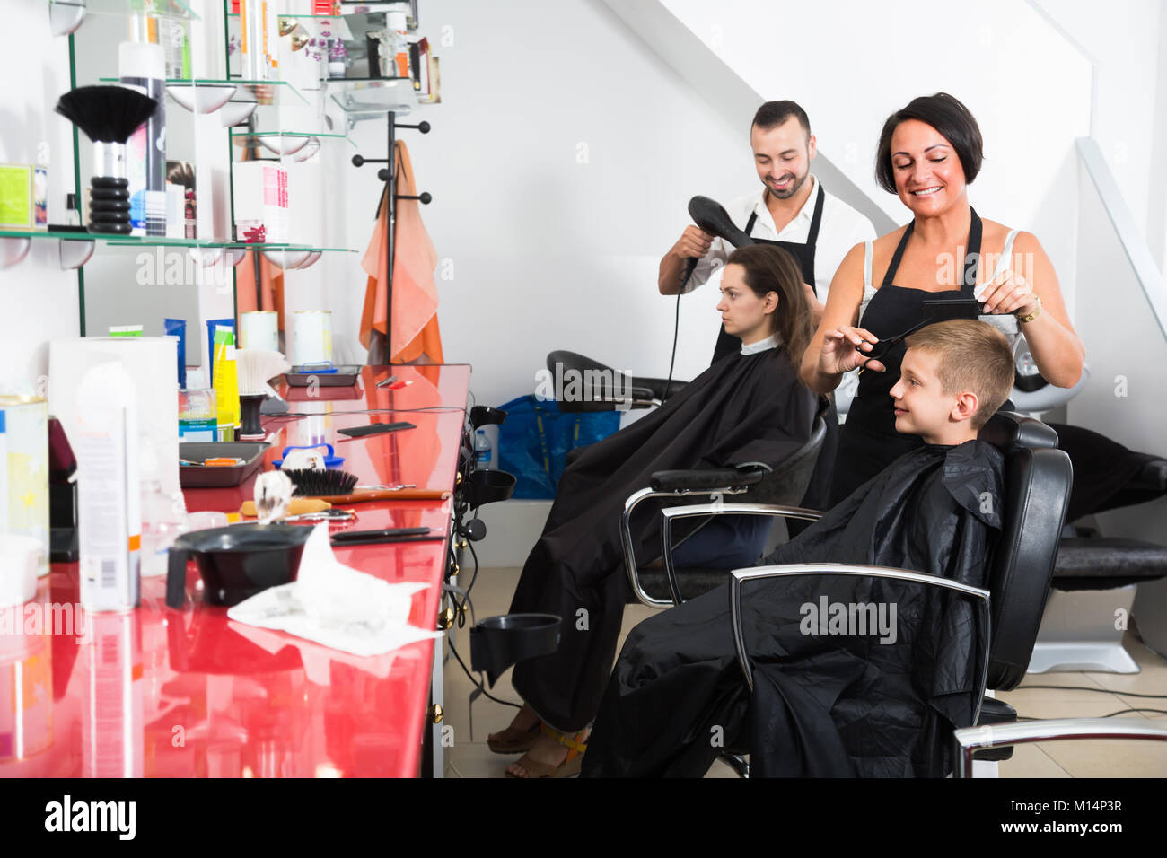 cheerful woman hairdresser cutting hair of boy in crowded beauty salon ...
