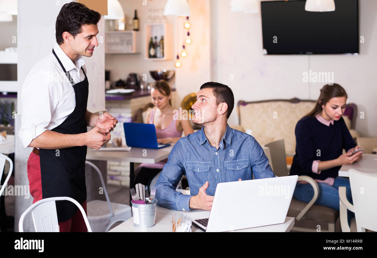 Portrait of polite cafe waiter standing at table and talking to guests ...