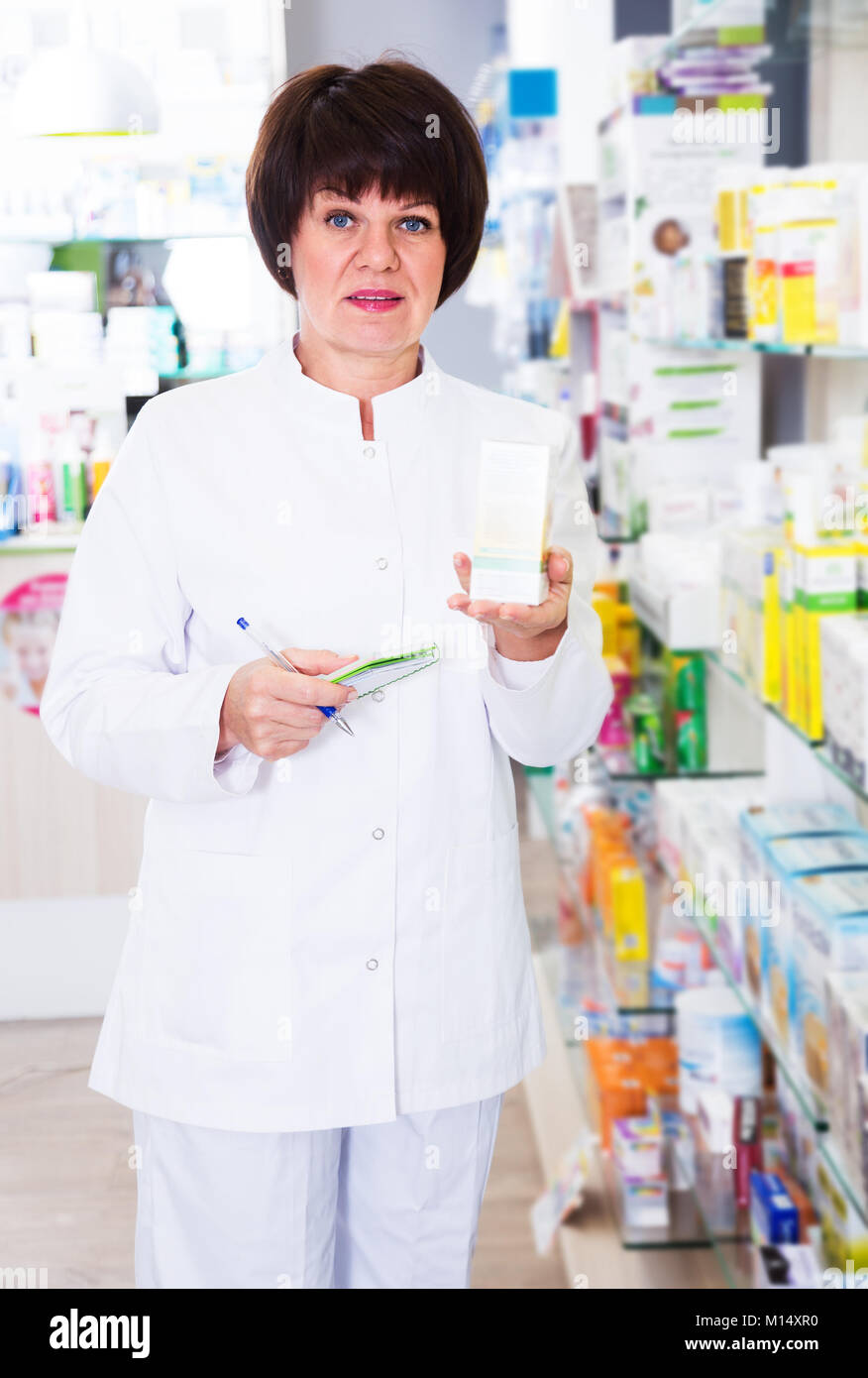 Positive woman druggist wearing white uniform standing among shelves in ...