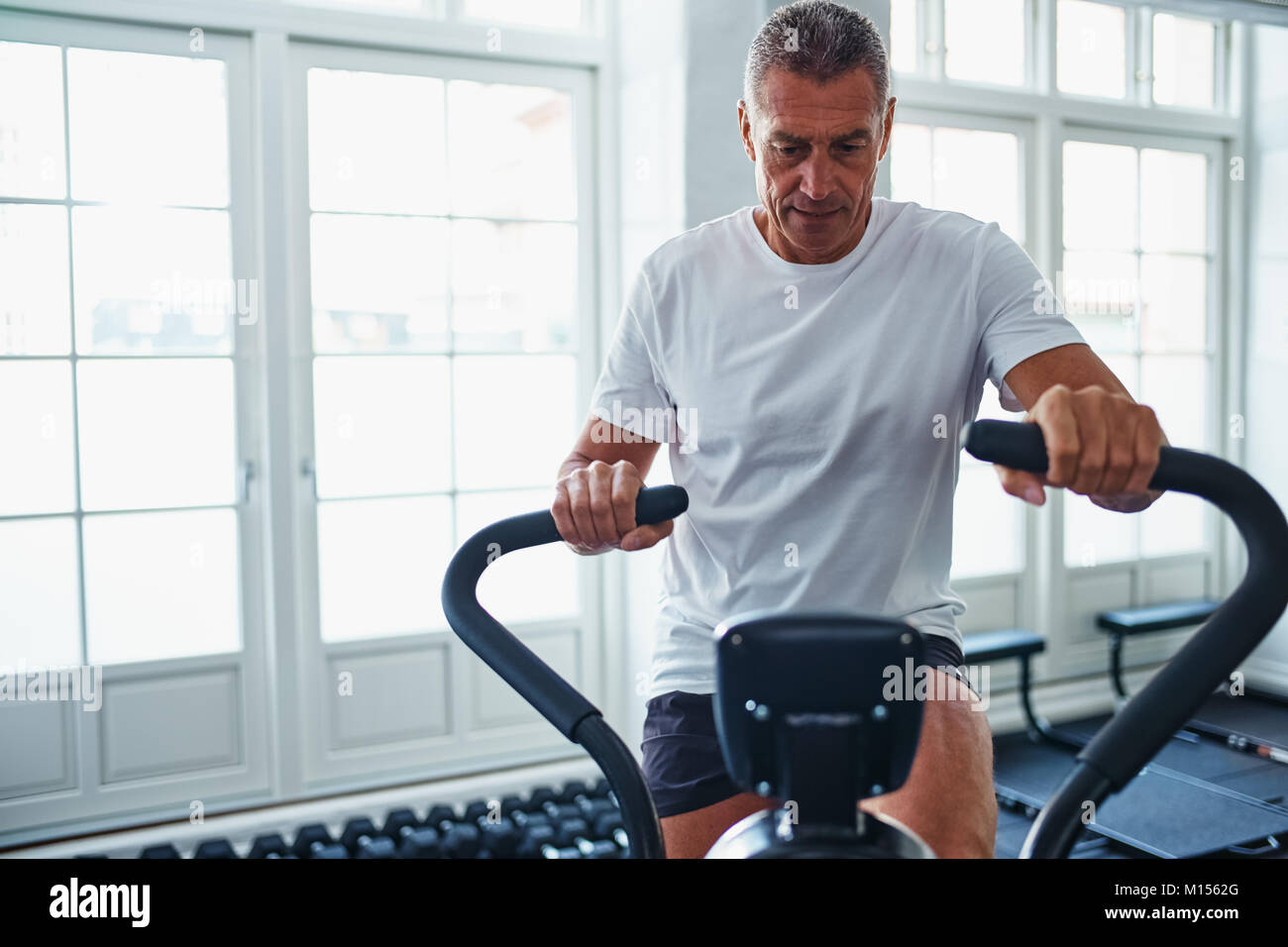 Focused mature man riding a stationary bike while working out alone at ...