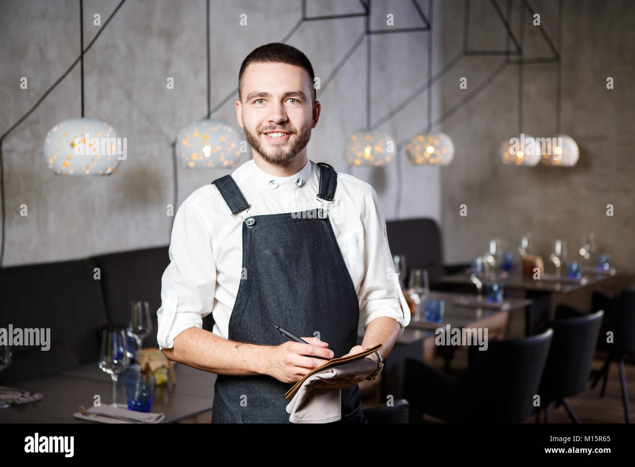A young, smiling waiter in a restaurant, standing next to the tables ...