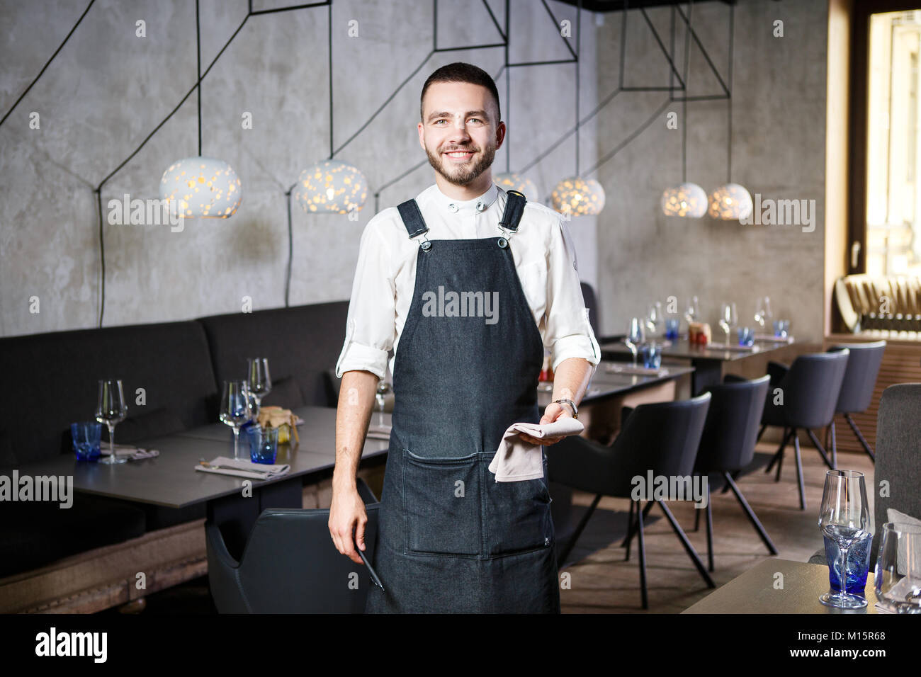 A young, smiling waiter in a restaurant, standing next to the tables ...