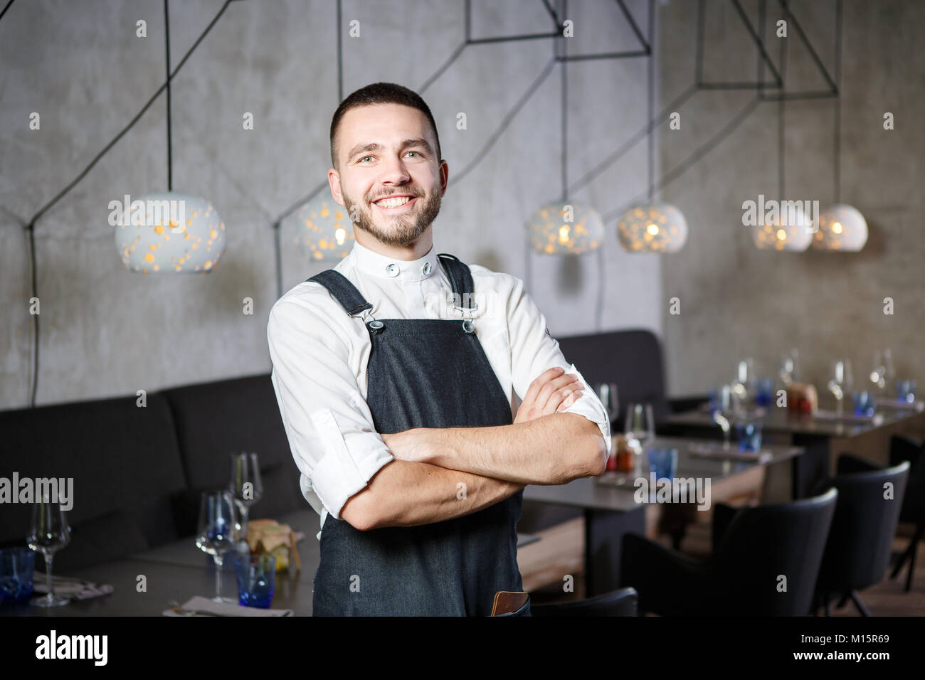 A young, smiling waiter in a restaurant, standing next to the tables ...