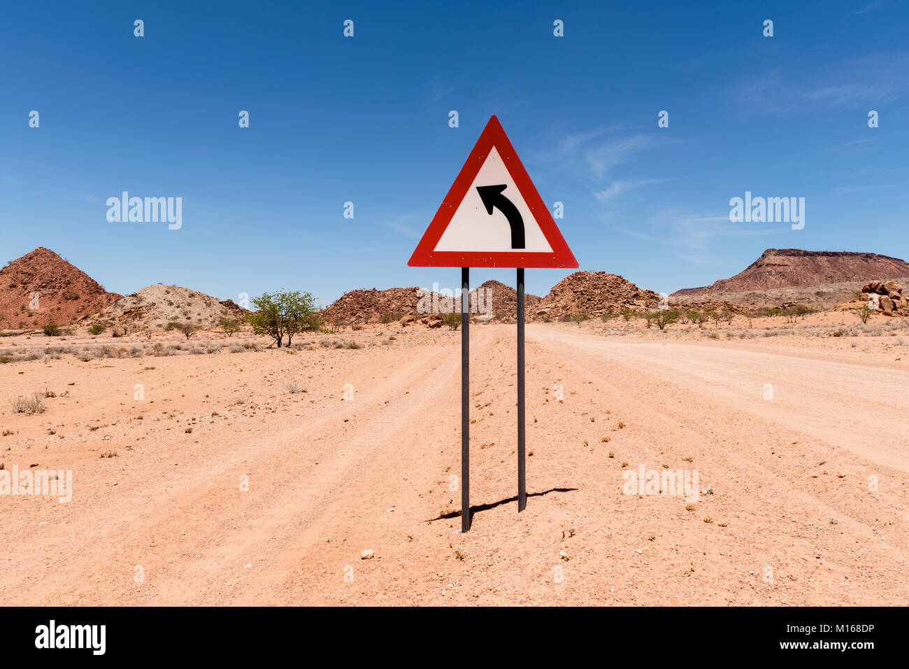 A road sign indicating a left hand bend, by the side of a gravel road ...