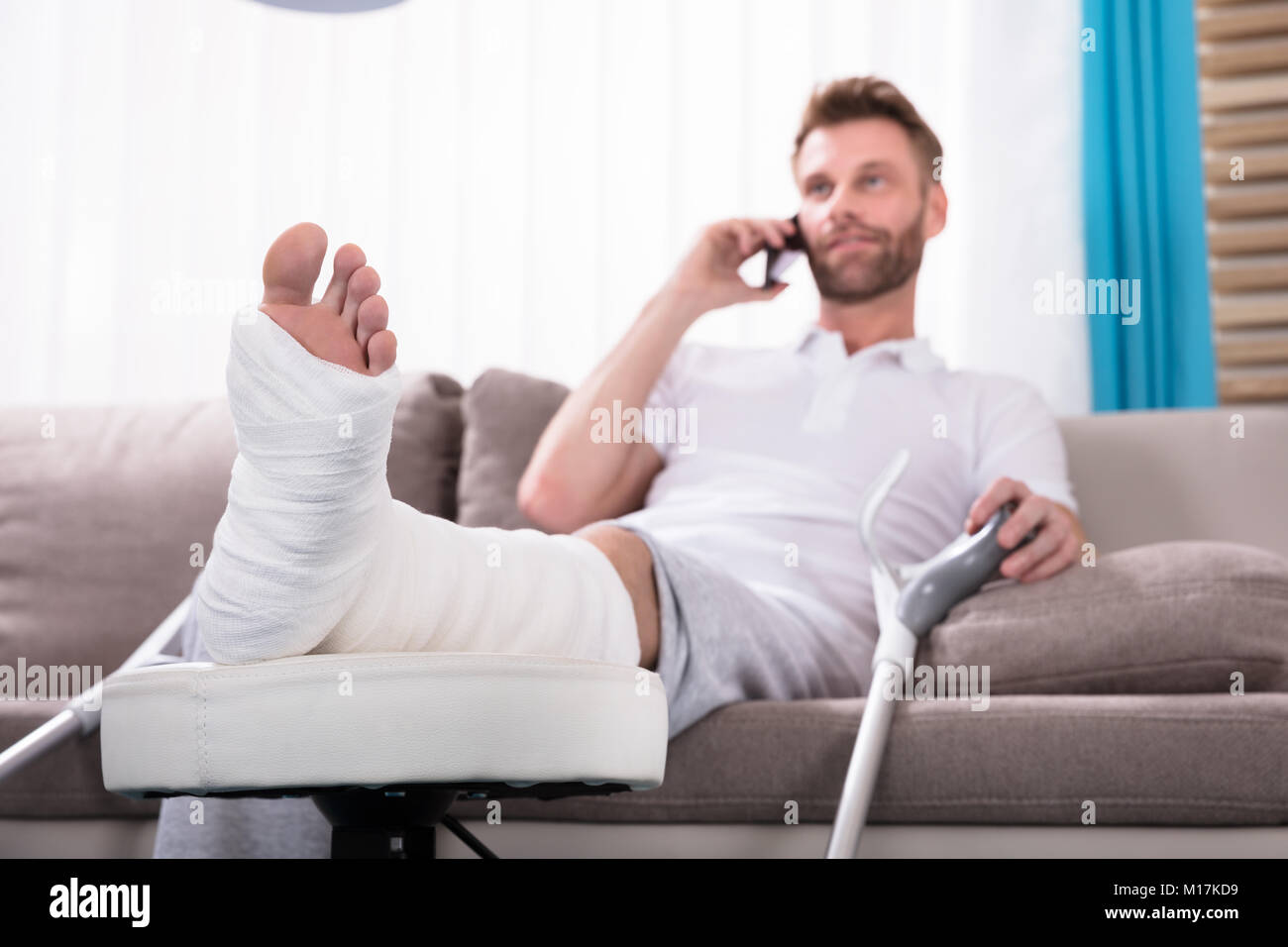 Happy Young Man With Broken Leg Sitting On Sofa Talking On Smartphone ...
