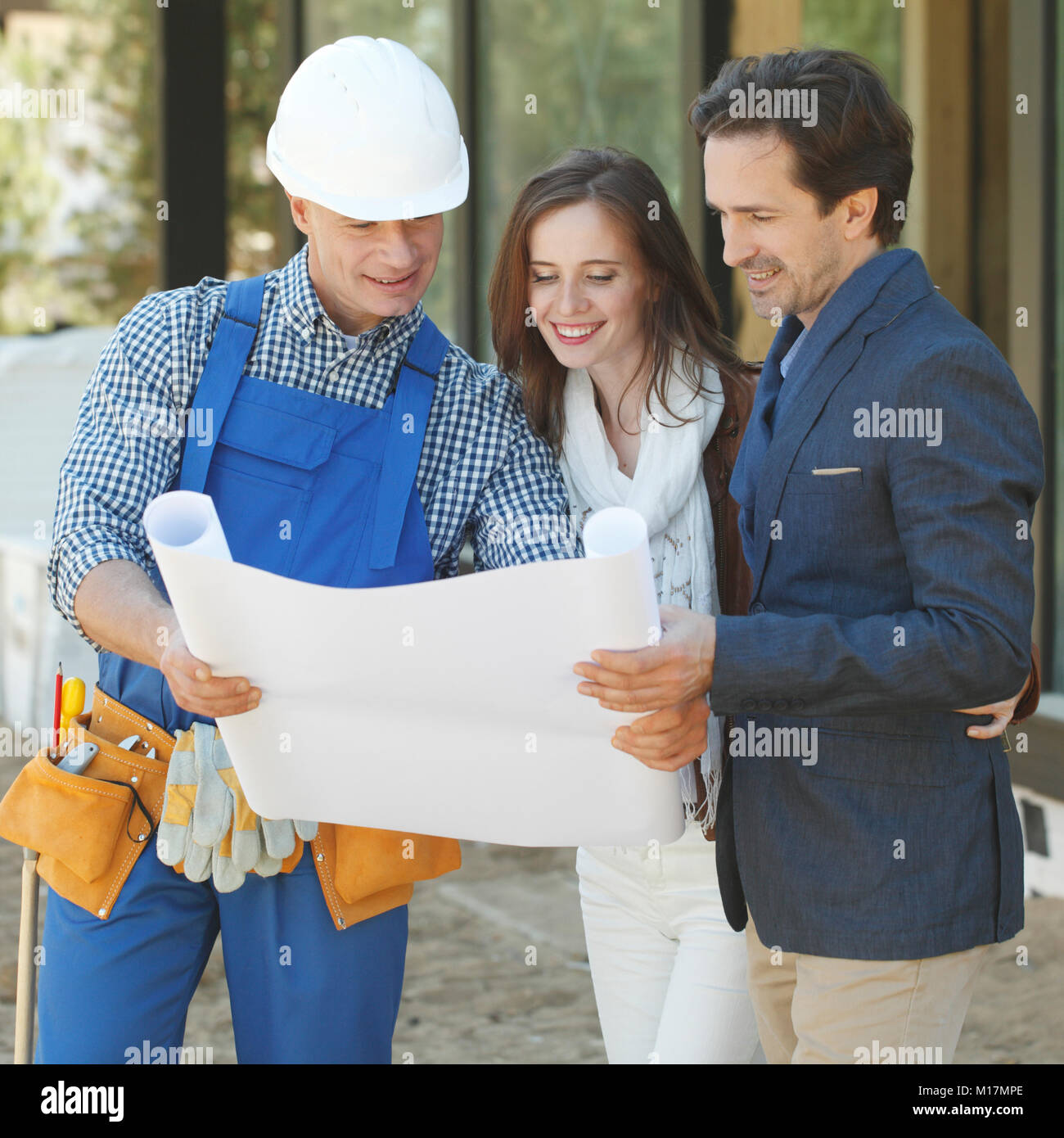 Worker shows house design plans to a young couple at construction site ...