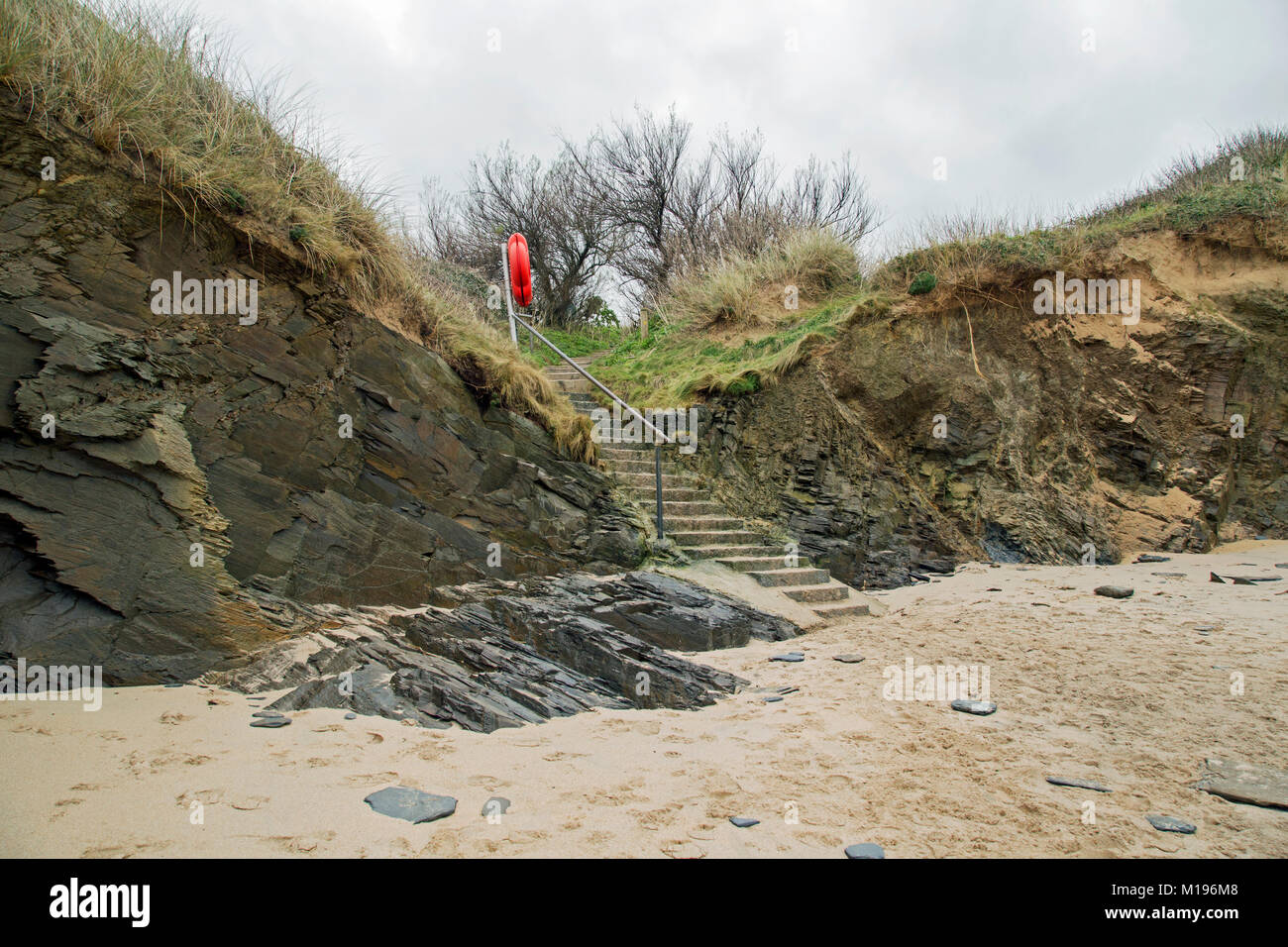 A view of a rocky Cornish beach with steps to a red life preserver belt ...
