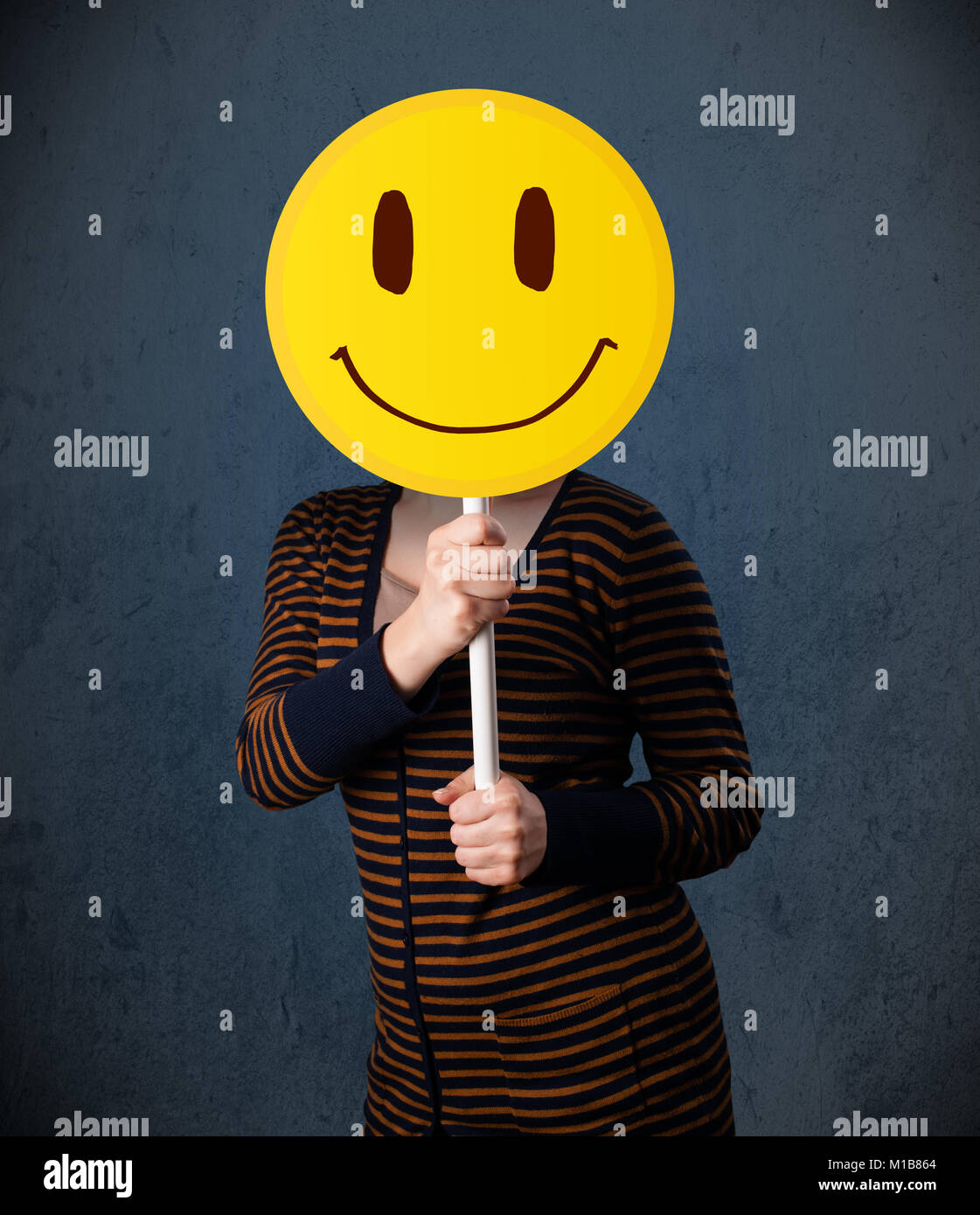 Young lady holding a yellow smiley face emoticon in front of her head ...