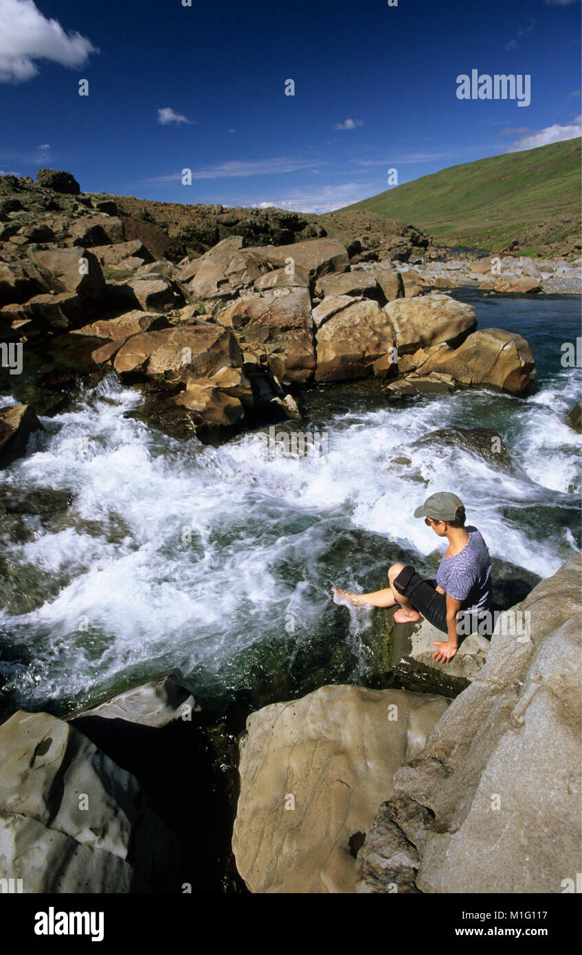 Iceland. near Borgarnes. Woman on rock by waterfall, dipping feet in ...