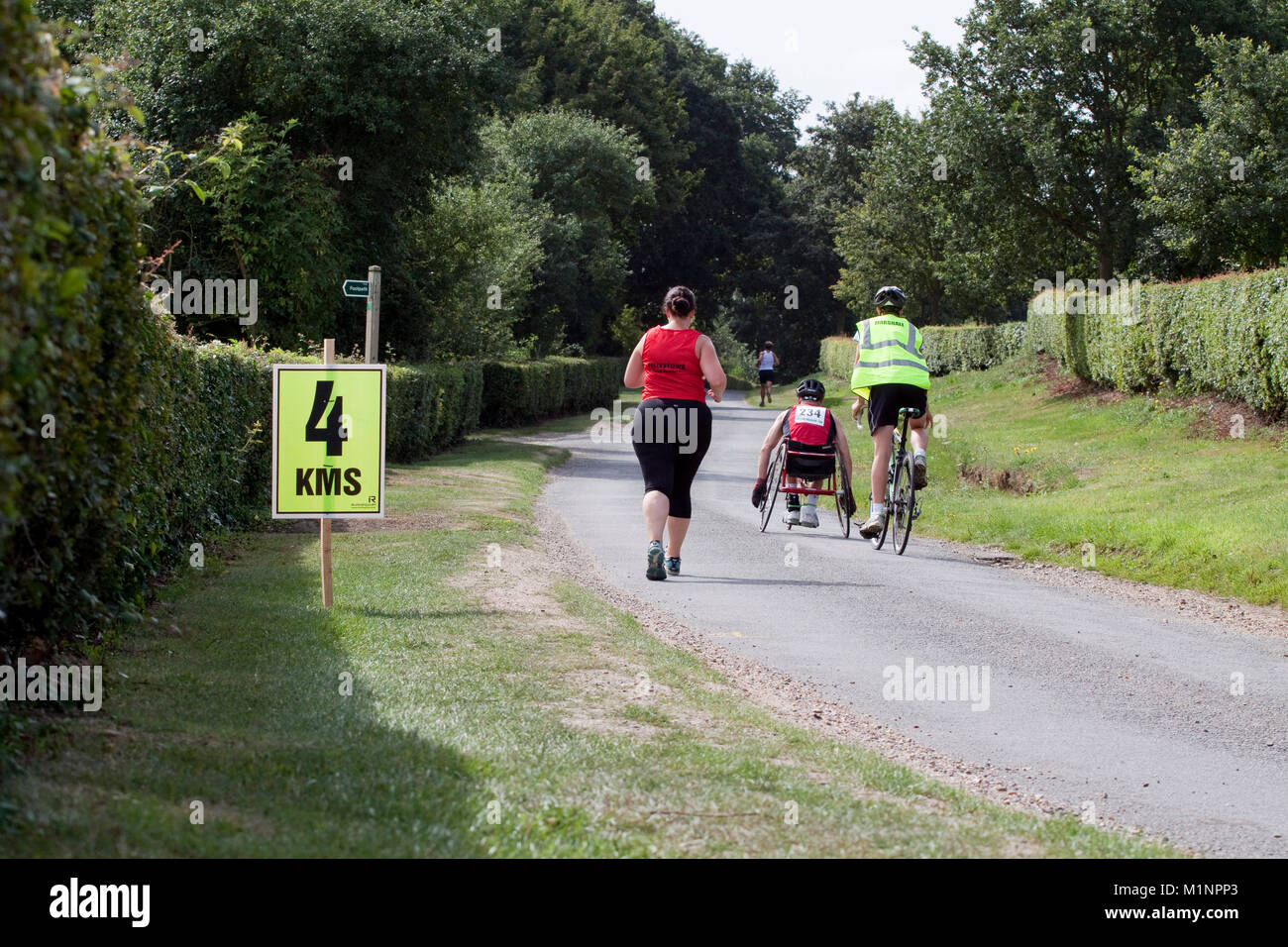 Wheelchair athlete with cycle marshal and woman runner pass 4 km sign ...
