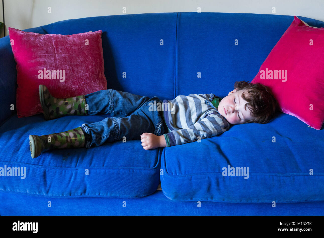 A young boy is fast asleep on a blue sofa in his clothes and wellies ...
