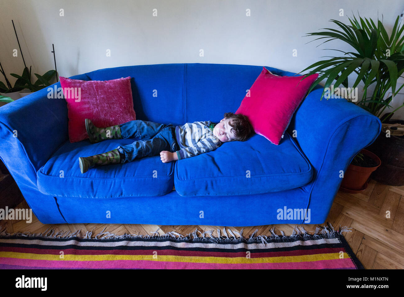 A young boy is fast asleep on a blue sofa in his clothes and wellies ...