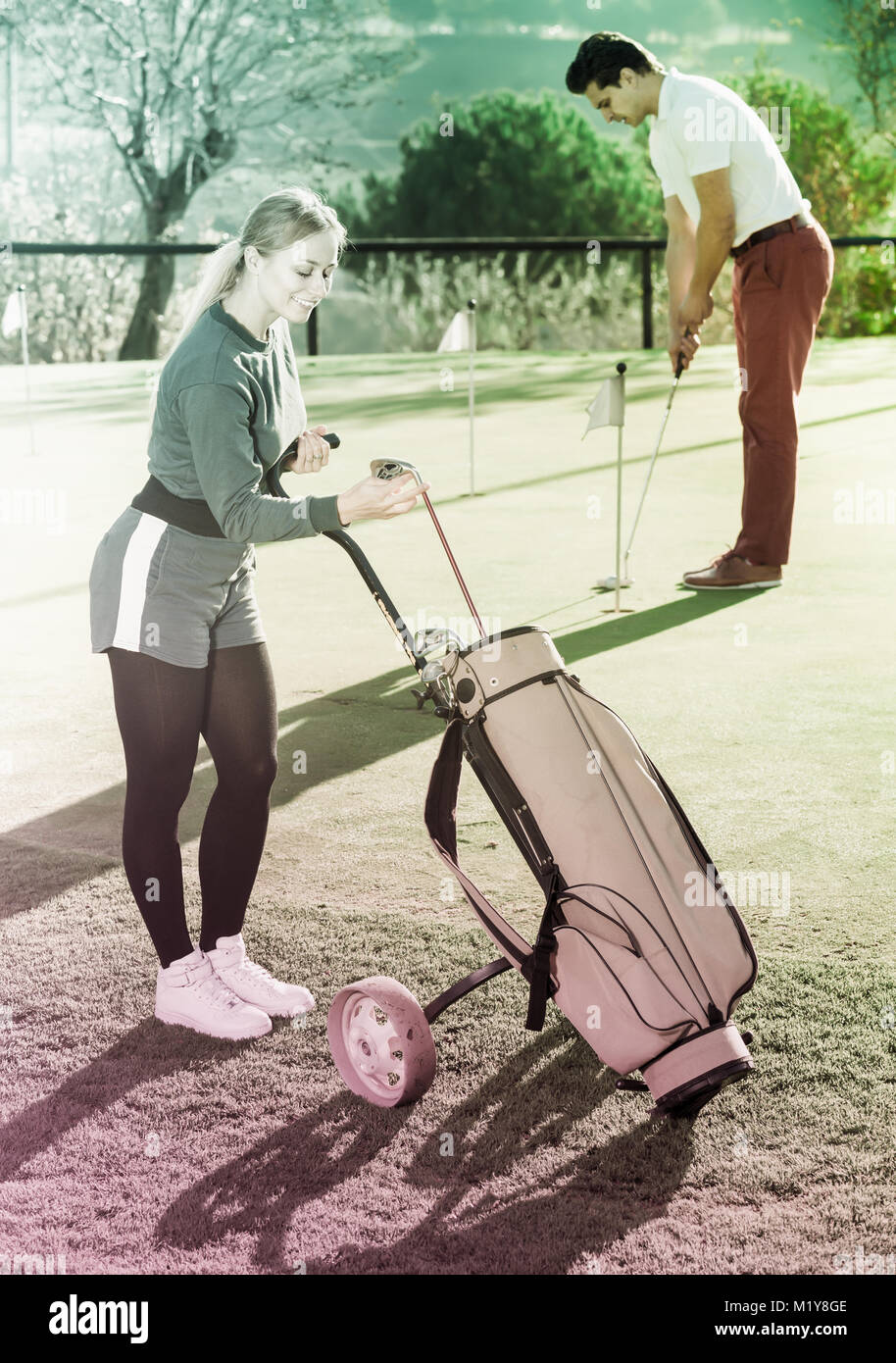 Female golfer examining clubs while her male golf partner hitting ball ...