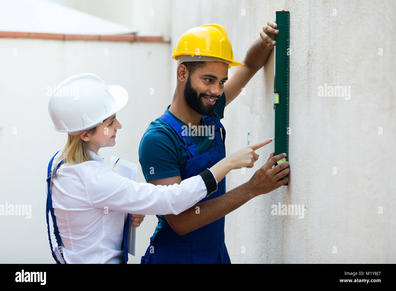 Smiling blonde female engineer and black specialist checking wall plane ...