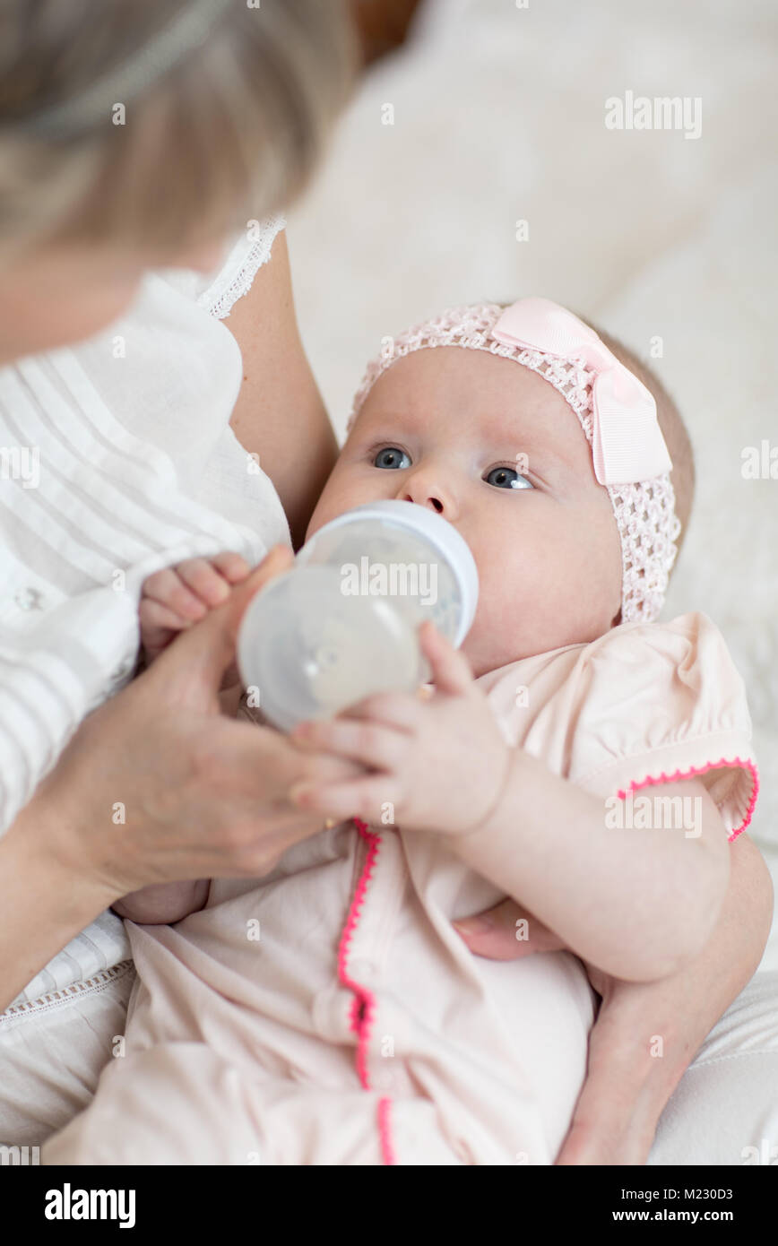 Mother giving baby girl nutrition from the bottle sitting on the bed ...