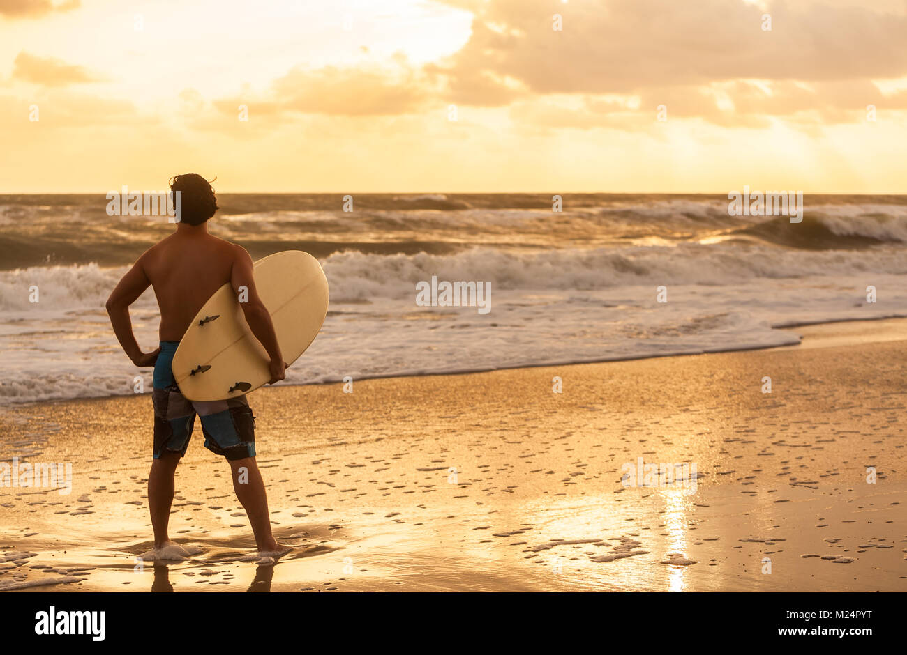 Rear view of young man male surfer with white surfboard looking at surf ...