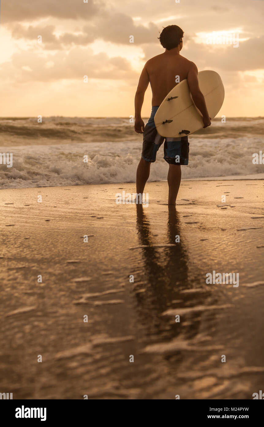Rear view of young man male surfer with white surfboard looking at surf ...