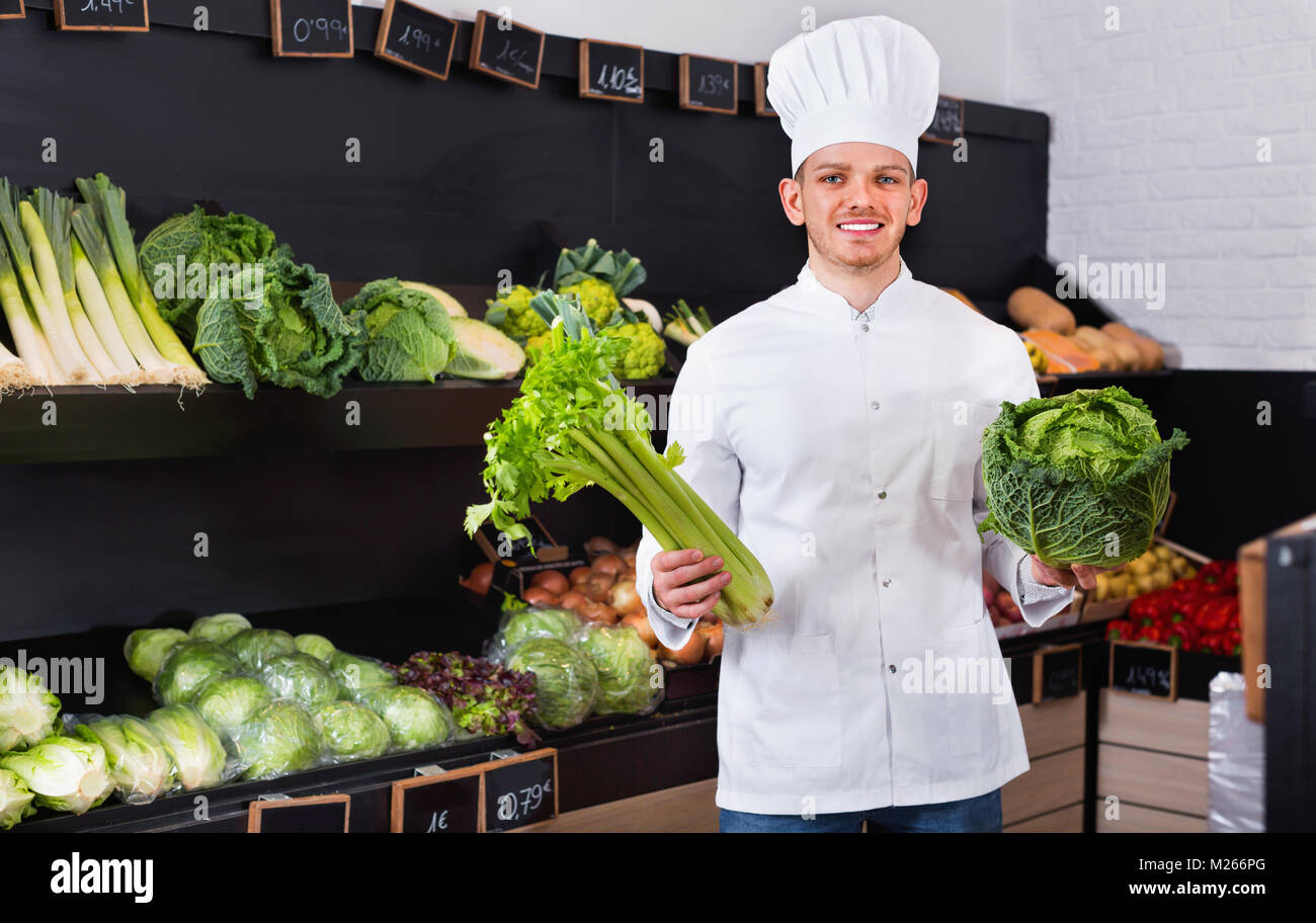Young satisfied smiling male cook choosing vegetables in grocery shop ...