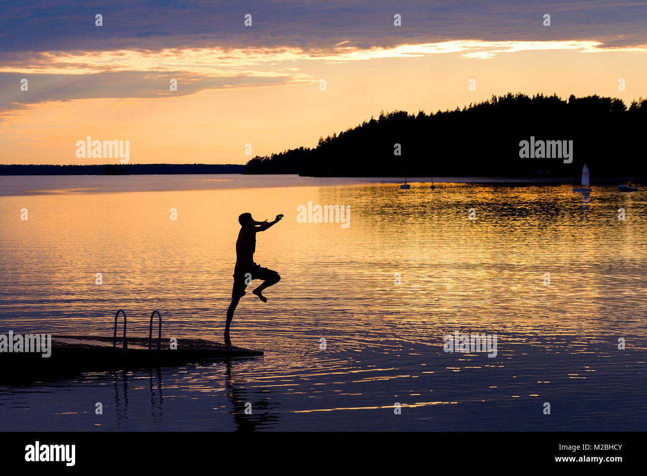 Silhouette of young man jumping from a pontoon into lake at sunset ...