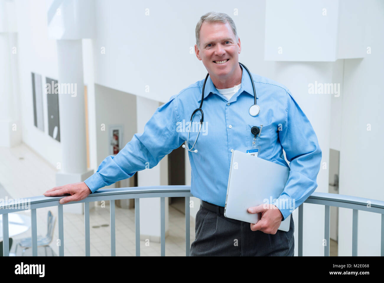 Portrait of smiling Caucasian doctor leaning on railing holding laptop ...