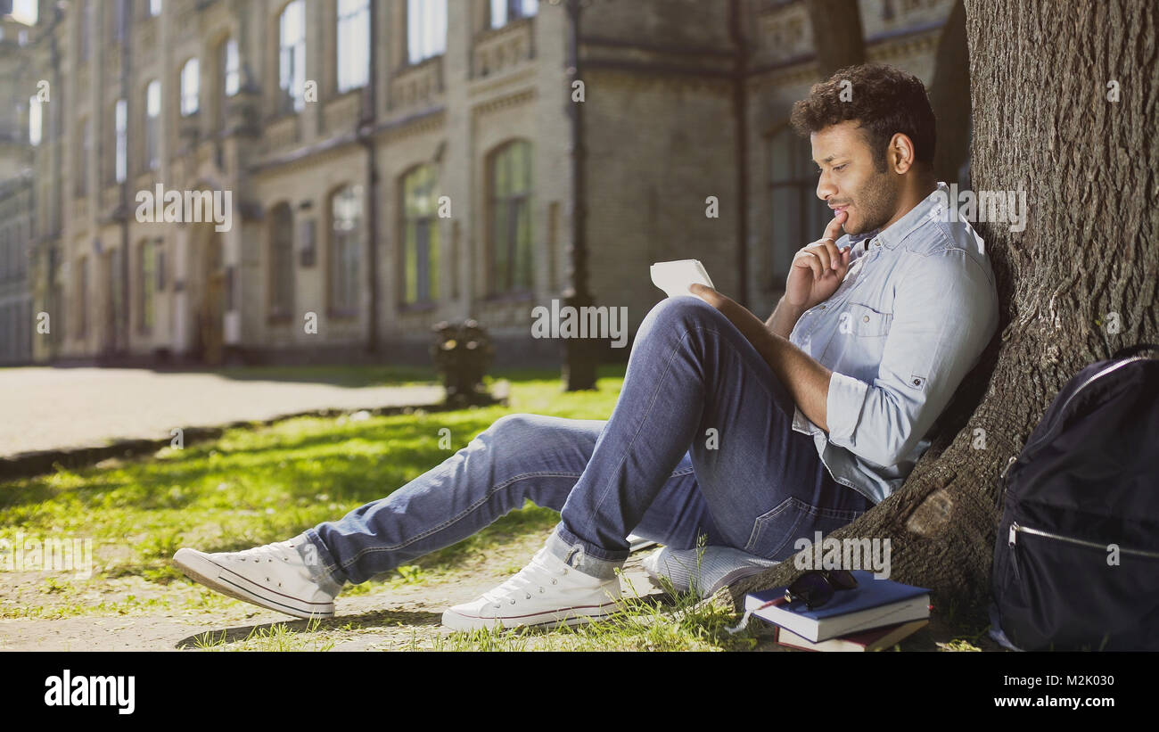 Multiracial young guy sitting with notebook under tree, thinking what ...