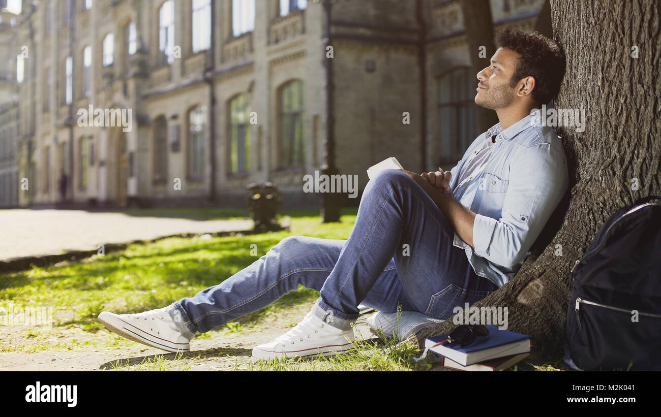 Dreamy young male sitting with notebook under tree, thinking what to ...